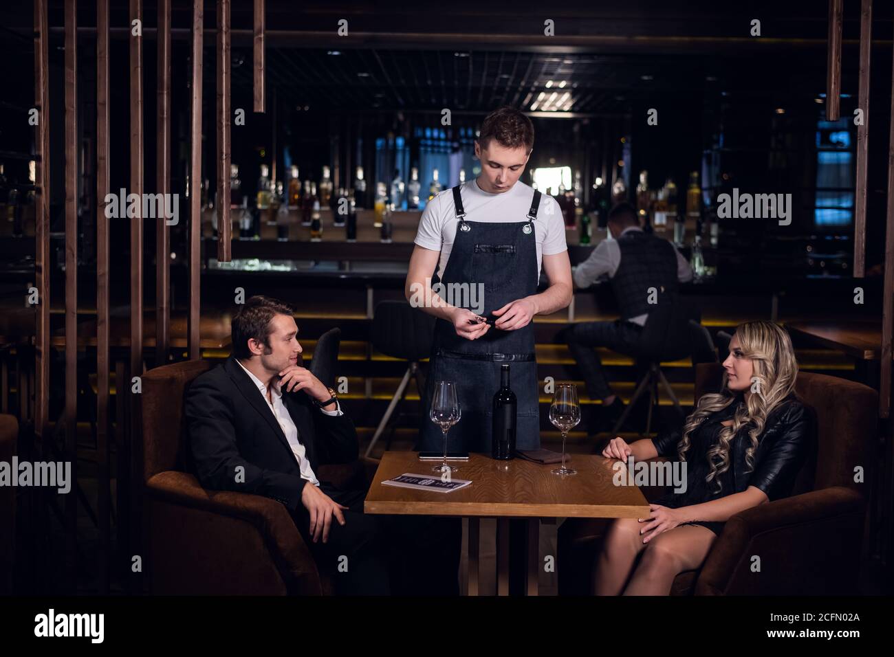 a young waiter guy opens a bottle of wine at the table in a restaurant ...