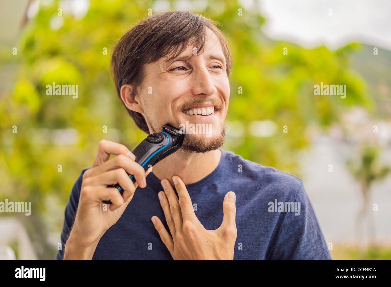 man is shaving off his beard with electric razor on the balcony during ...