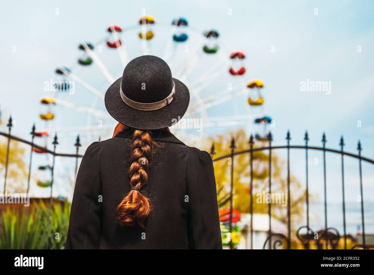 Back view of a young girl in hat standing in front of the ferris wheel ...