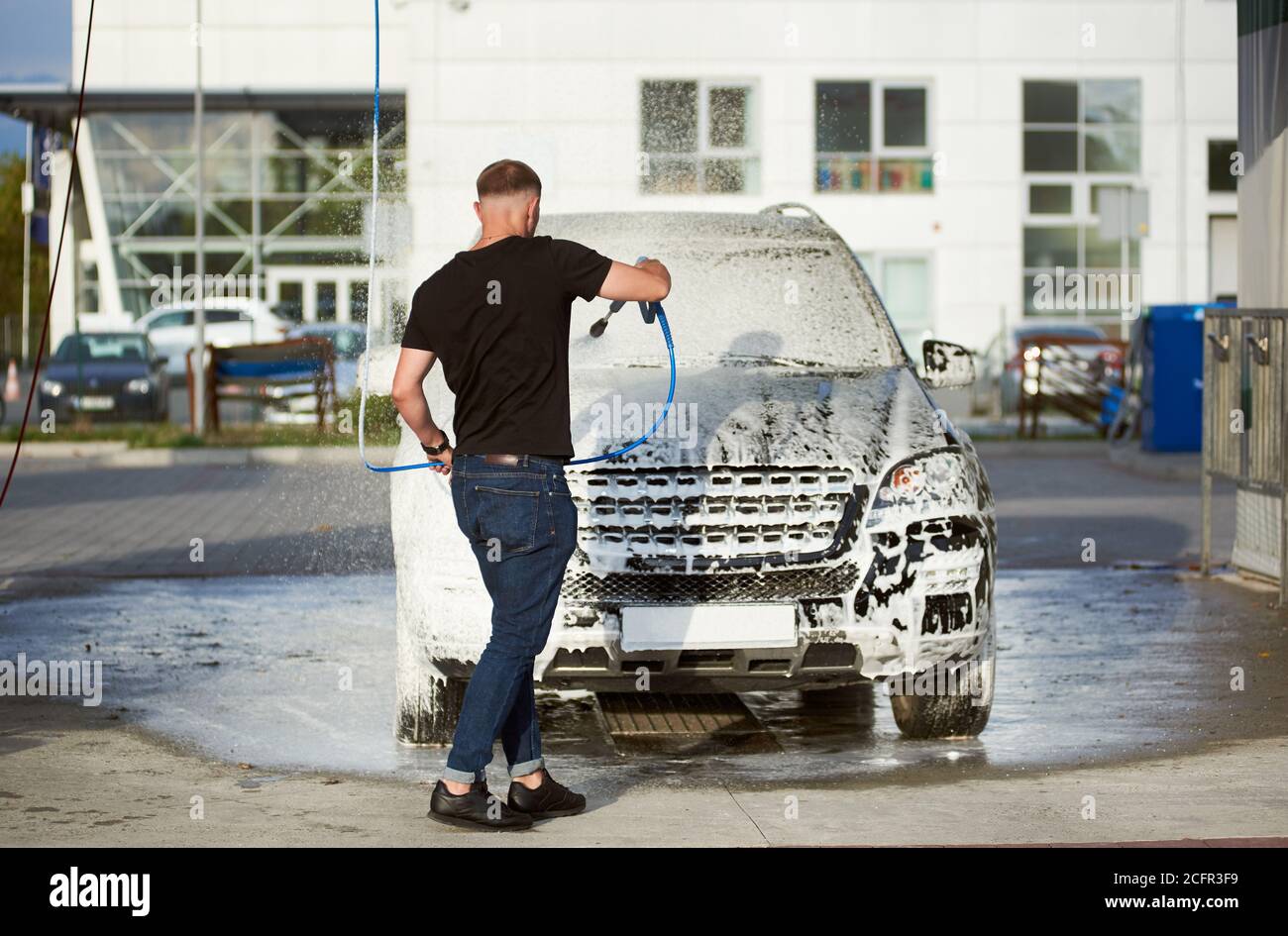Back view of a sporty man holding a hose with soap and washing his car ...