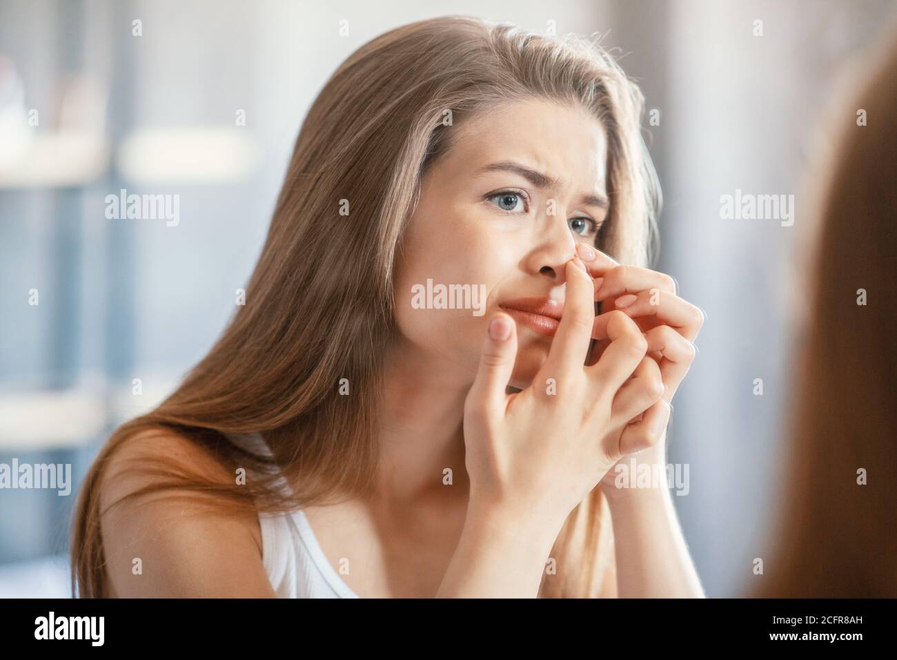 Displeased young woman squeezing acne on her nose while looking in ...