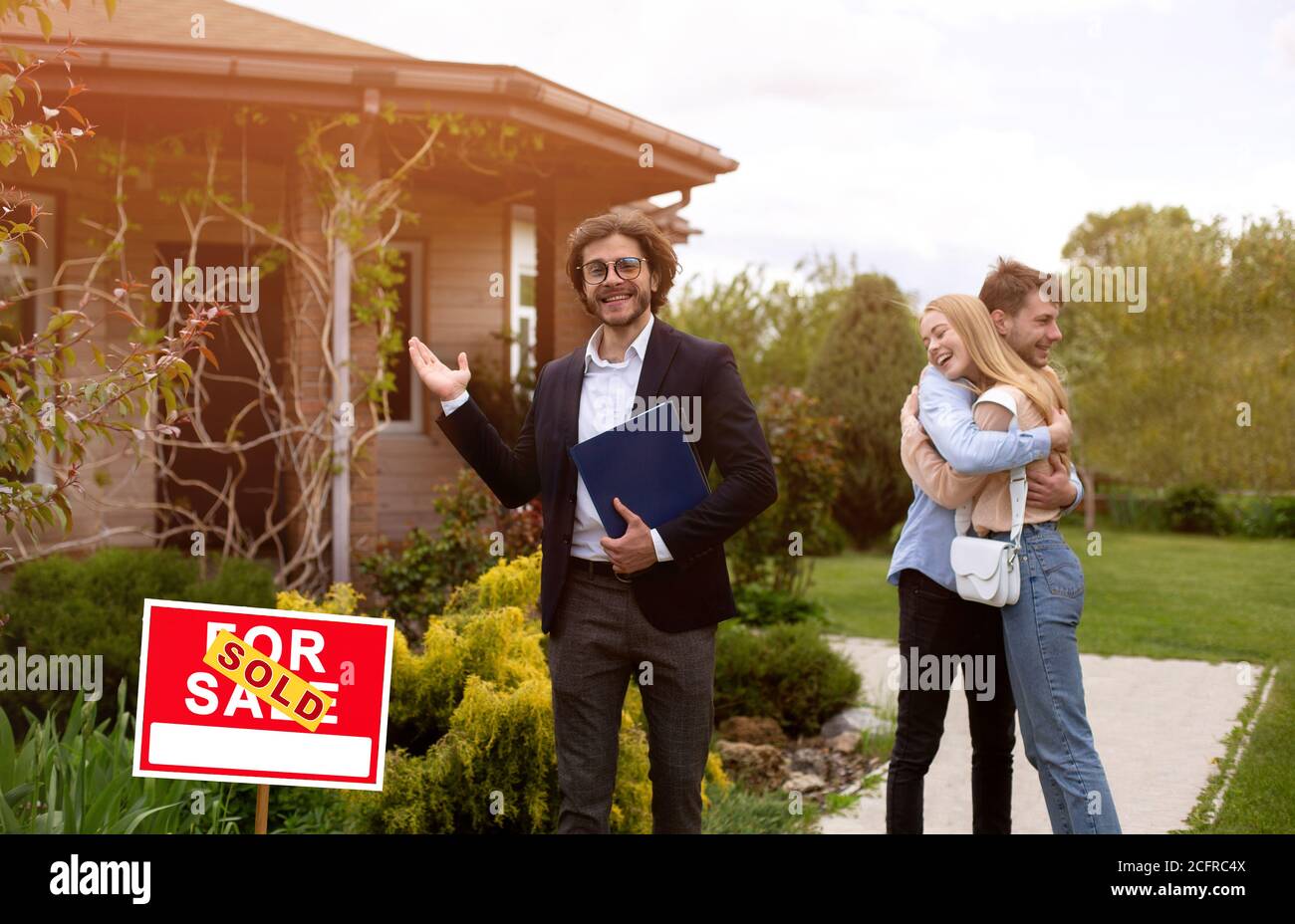 Smiling house agent and happy couple embracing in front yard of their ...