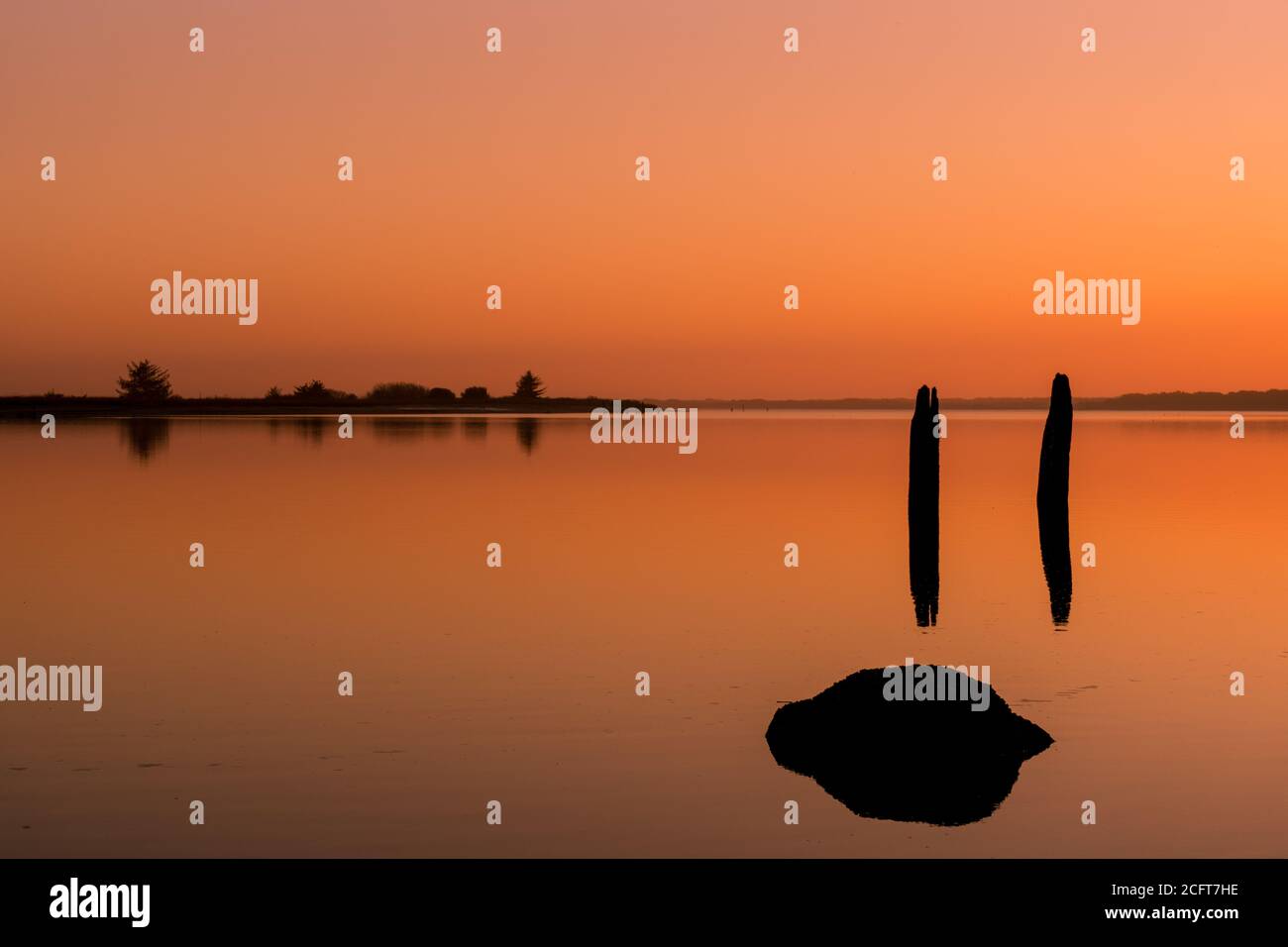 Very orange sunset with pilings and logs reflect in glassy river ...