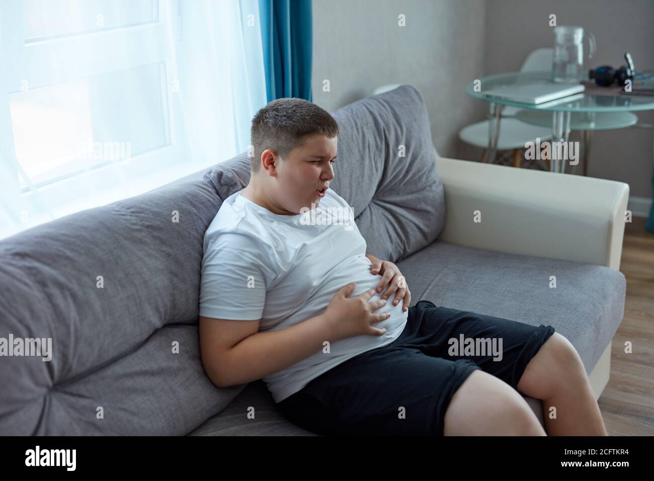 child boy has bloating after a fatty meal, he sits on sofa holding arm ...