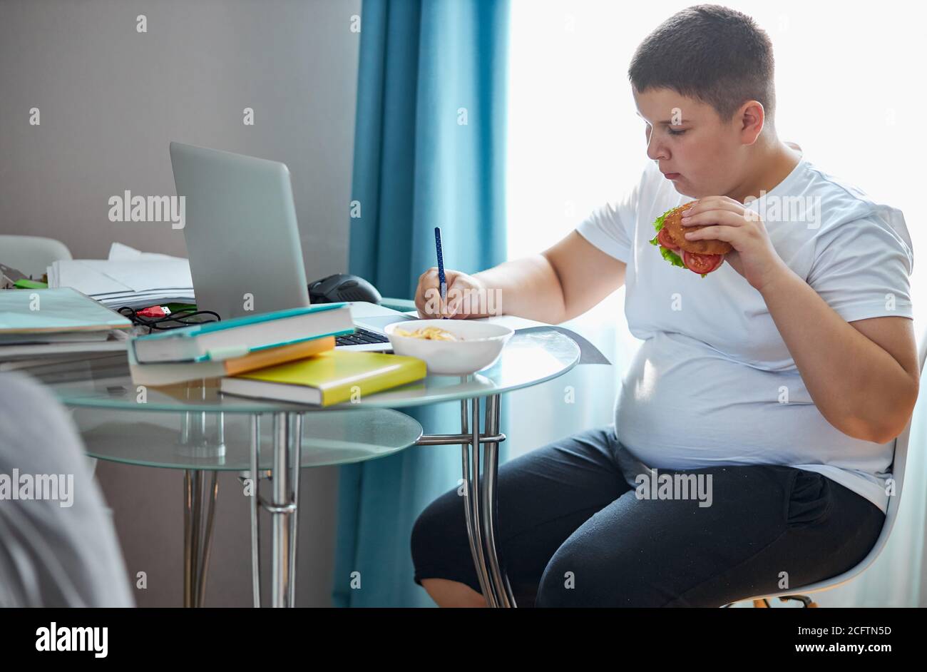 overweight school boy eat sandwich while doing homework, sits at table ...