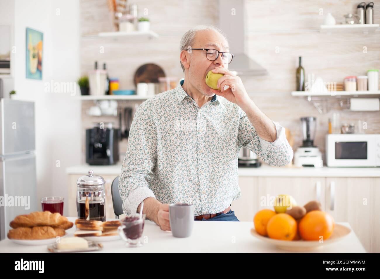 Elderly retired person biting apple during breakfast in mdoern cozy ...