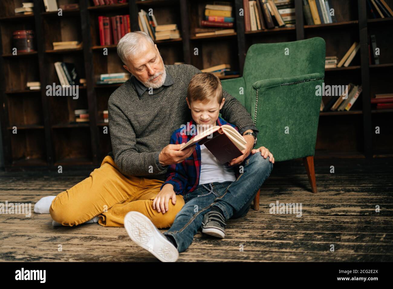 Senior grandfather and grandson reading book together sitting on floor ...
