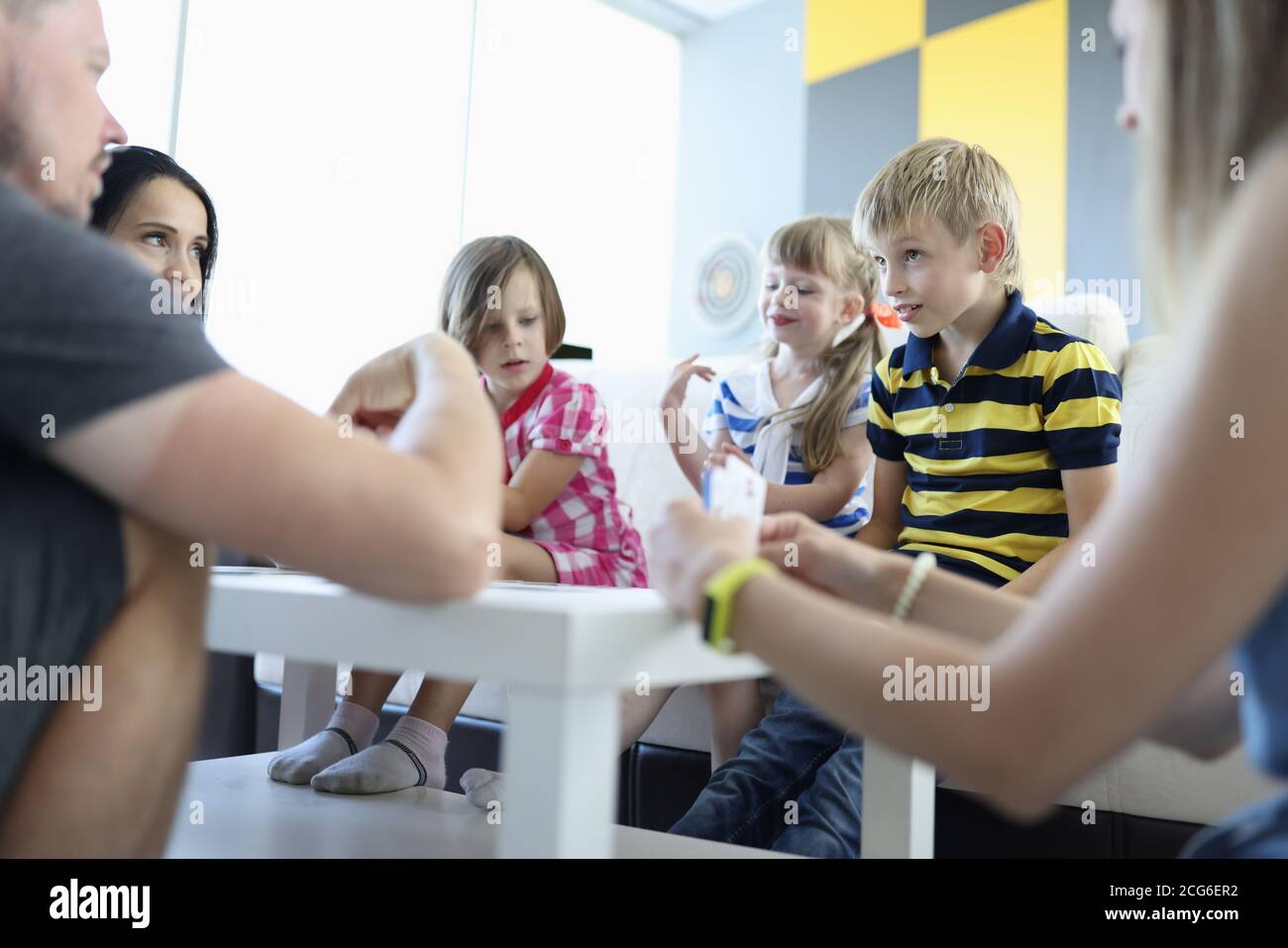 Adults and children are sitting around table on which playing cards are ...