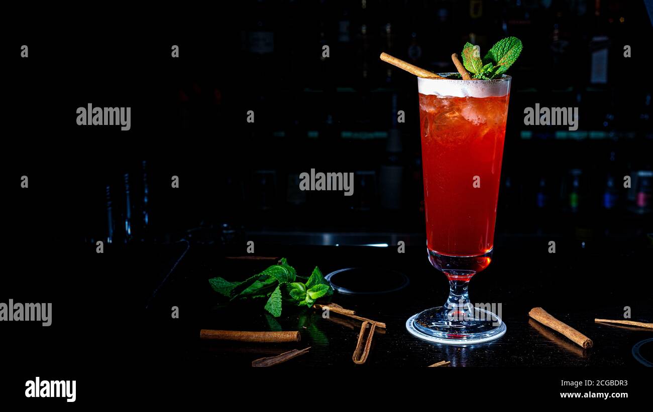 A red cocktail in a tall glass stands on the counter of a dark bar ...