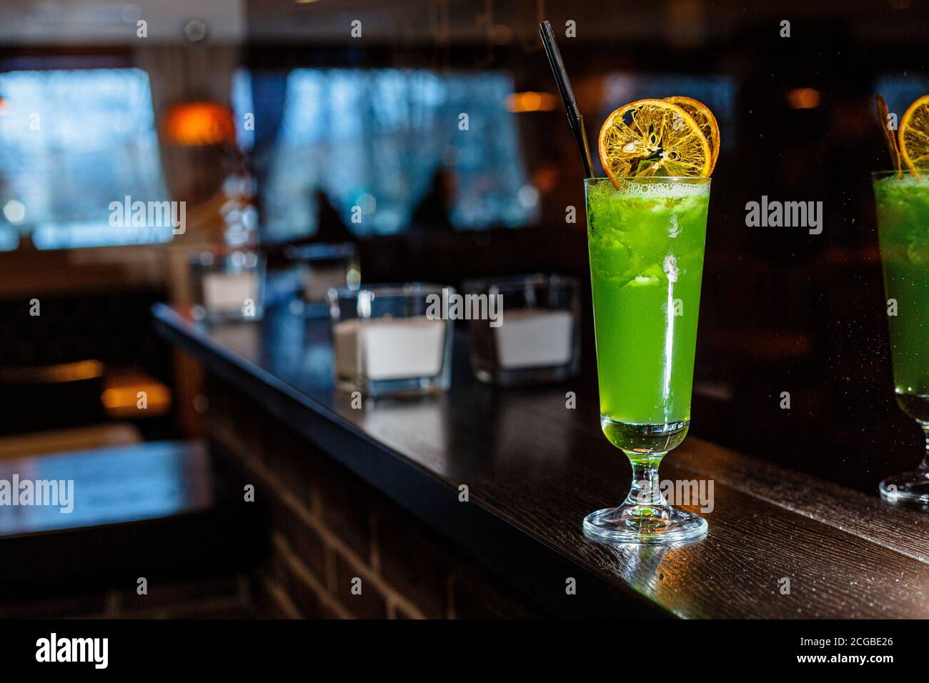 A green cocktail in a tall glass stands on the counter of a dark bar ...
