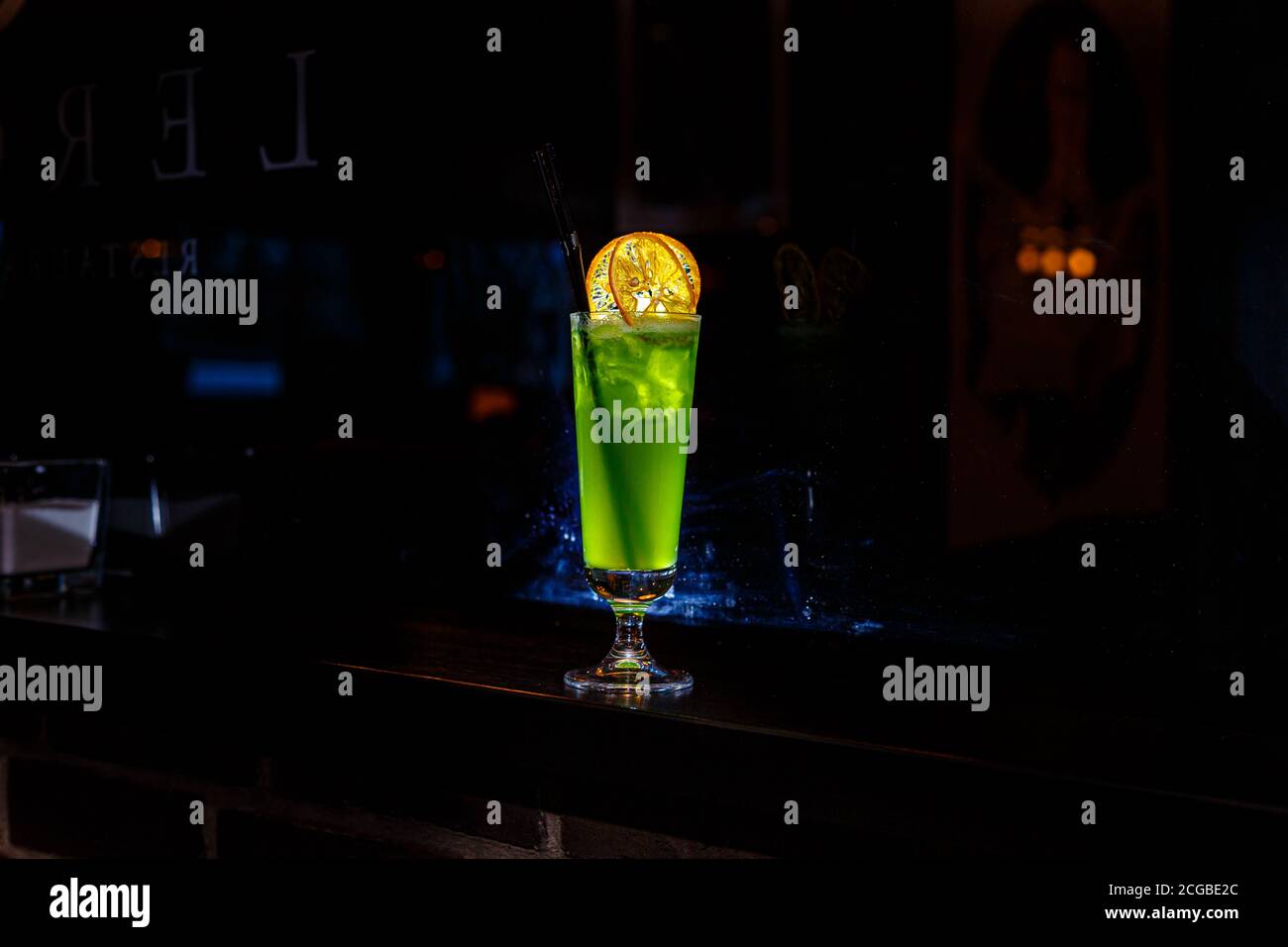A green cocktail in a tall glass stands on the counter of a dark bar ...