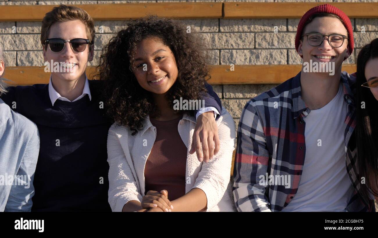 Five young adult friends sitting in a row on a bench smiling to Stock ...