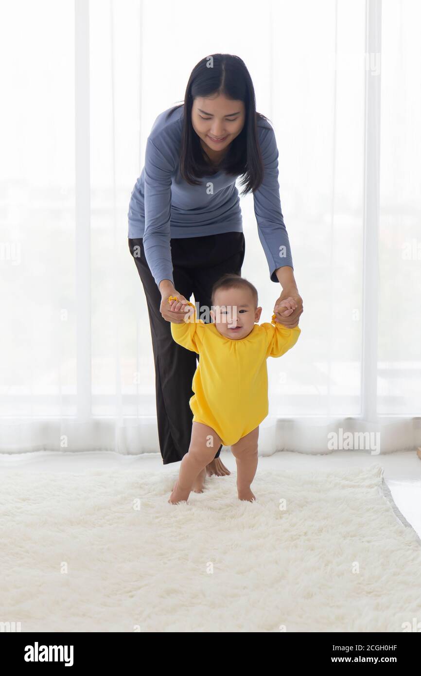 An Asian mother is helping to support her baby to learn to walk in a ...
