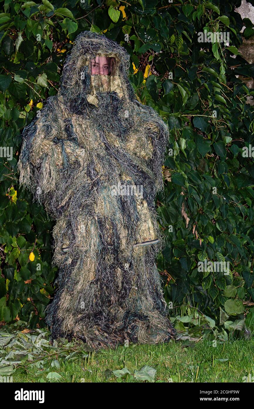 A person in camouflage stands almost invisible in front of a hedge ...