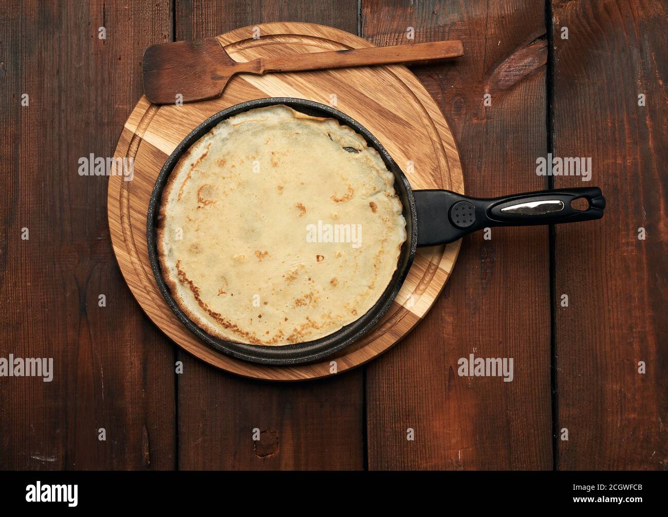 baked round big pancake in a black frying pan on a brown wooden table, top view, carnival Stock ...