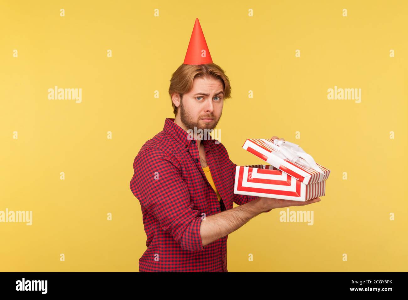 Unhappy sad guy in checkered shirt and party cone opening gift box ...