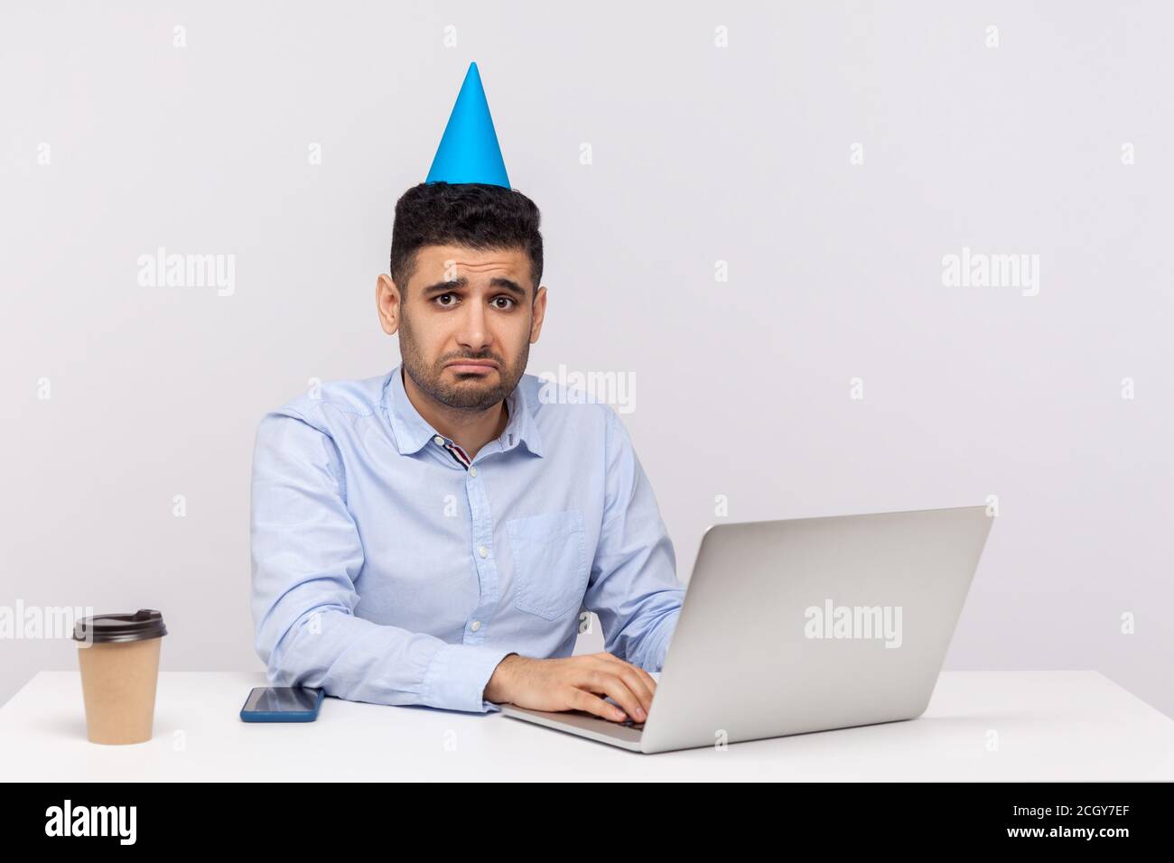 Unhappy employee guy with funny party cone on head sitting in office ...