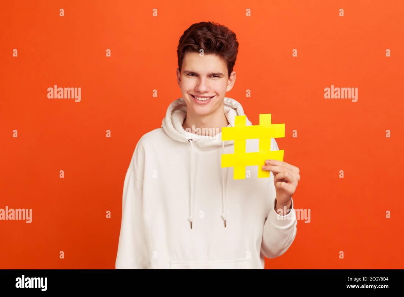 Handsome cheerful teenager in casual hoodie holding hashtag symbol with ...