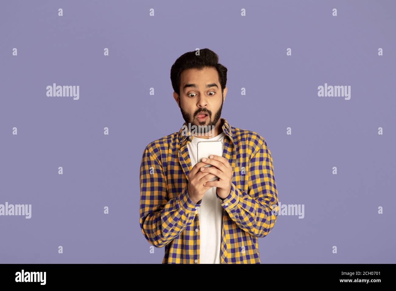 Handsome Indian man looking at his smartphone in shock on lilac ...