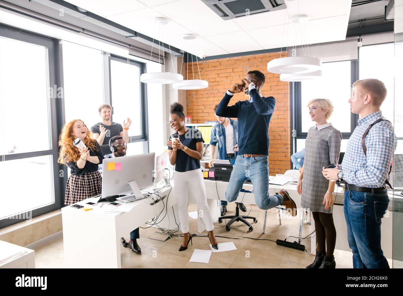 jumping African guy among laughing workers in the room with desks and ...