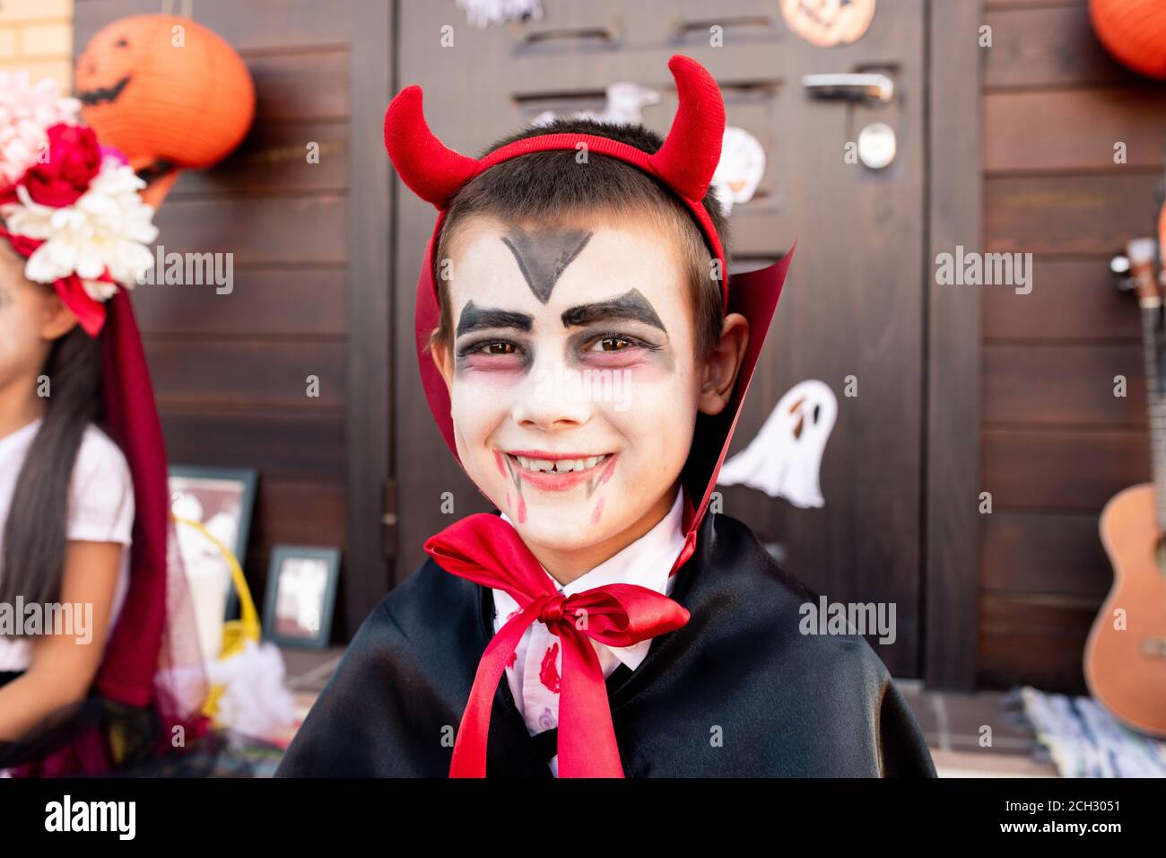Cheerful boy with painted face in costume of devil sitting in front of ...