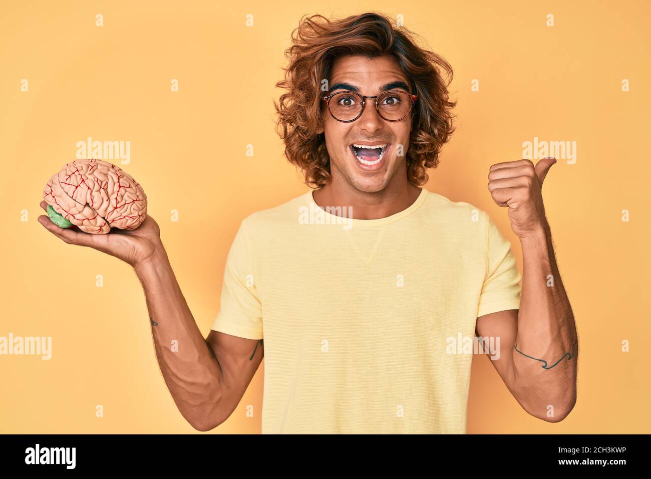 Young hispanic man holding brain pointing thumb up to the side smiling ...