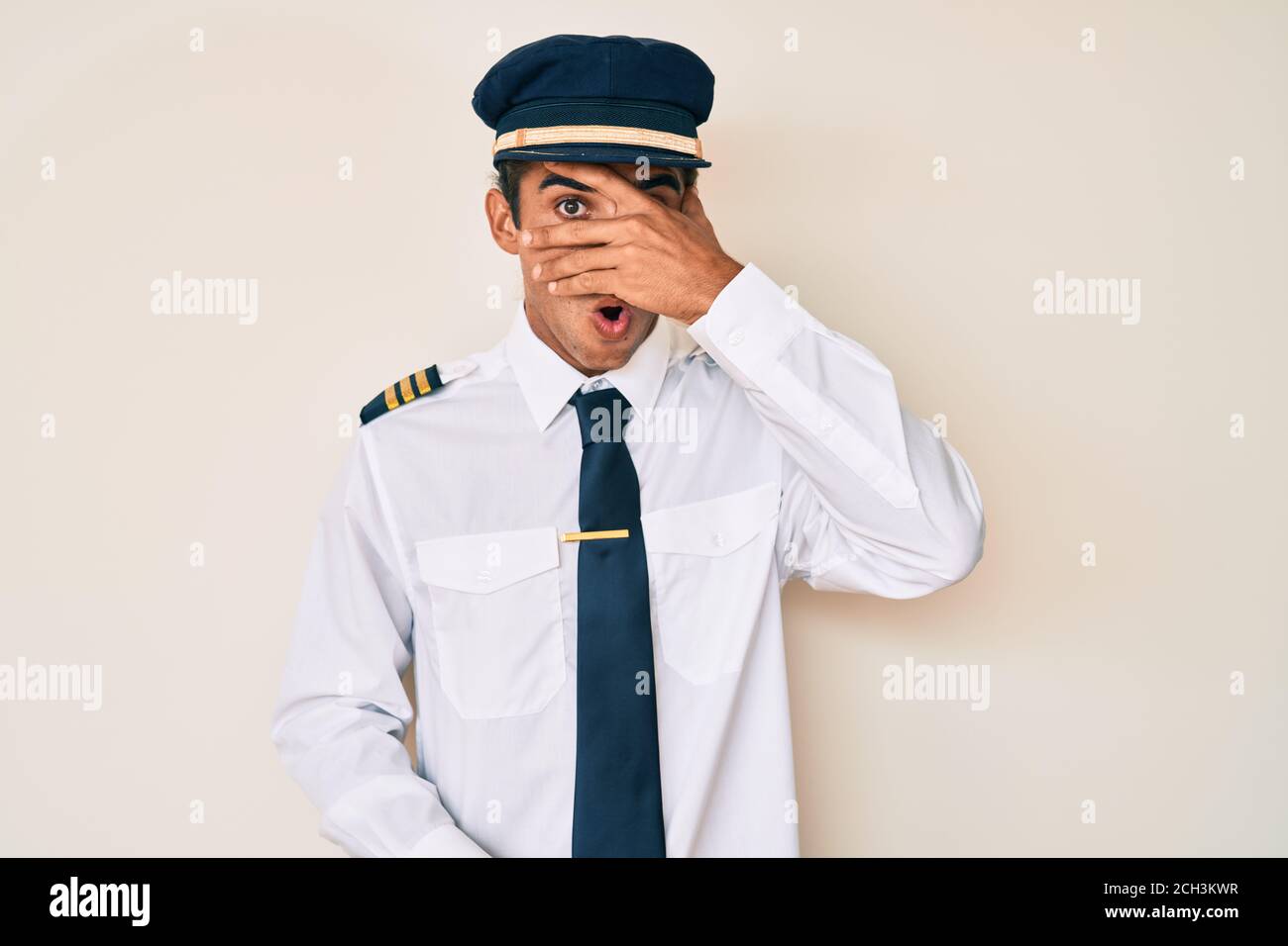 Young hispanic man wearing airplane pilot uniform peeking in shock ...