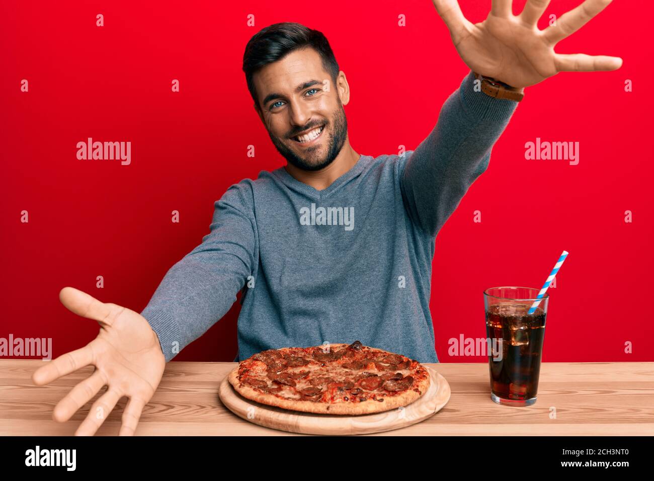 Handsome hispanic man eating tasty pepperoni pizza looking at the camera smiling with open arms ...