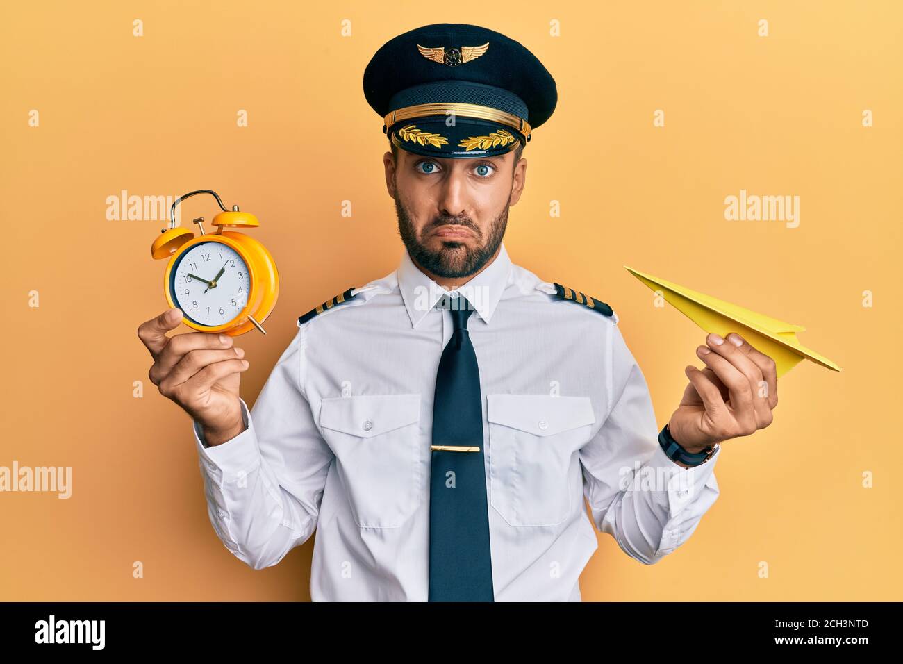 Handsome hispanic pilot man holding paper plane and alarm clock ...
