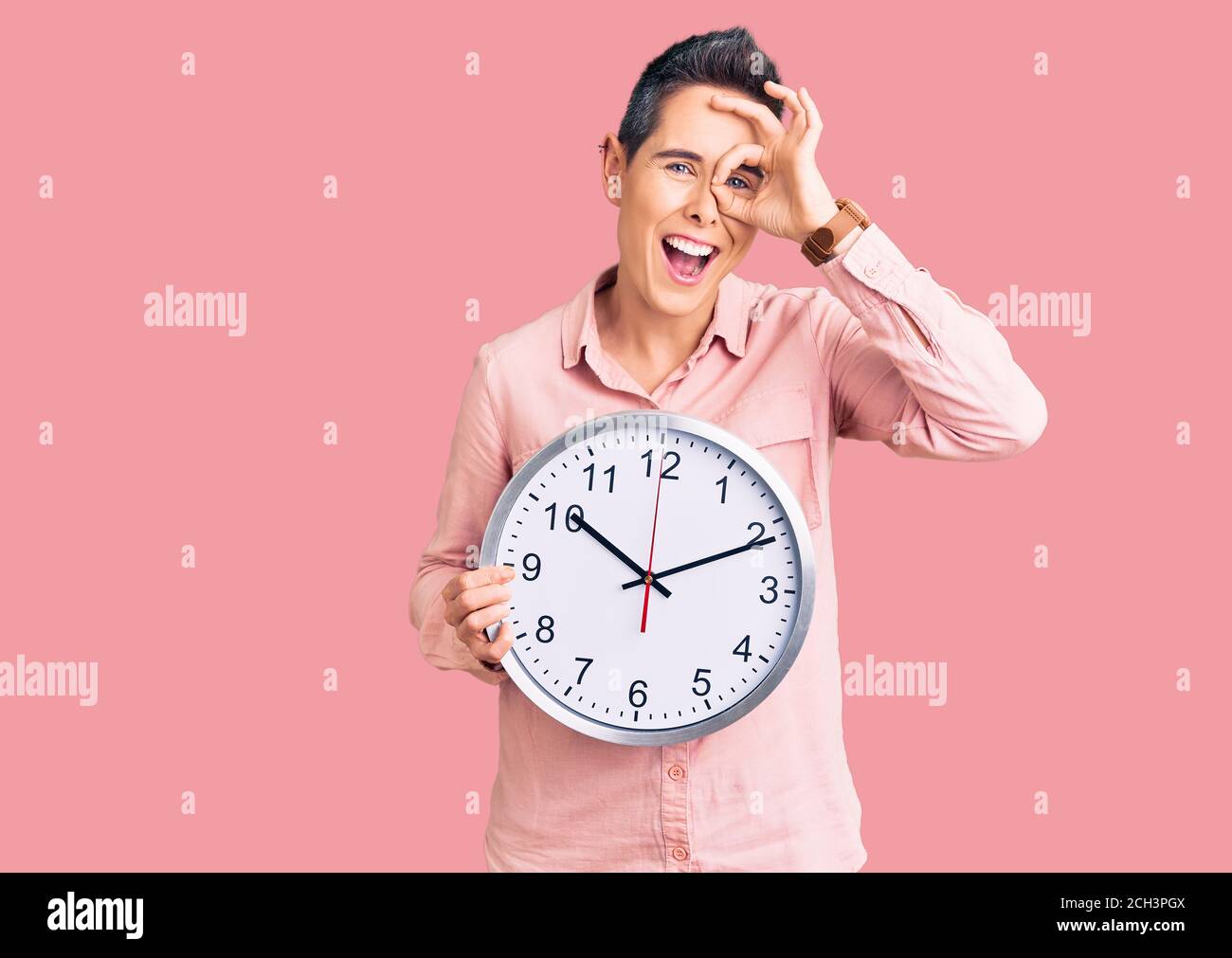 Young woman with short hair holding big clock smiling happy doing ok ...