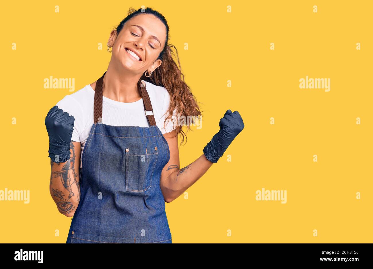 Young hispanic woman with tattoo wearing barber apron and gloves very ...