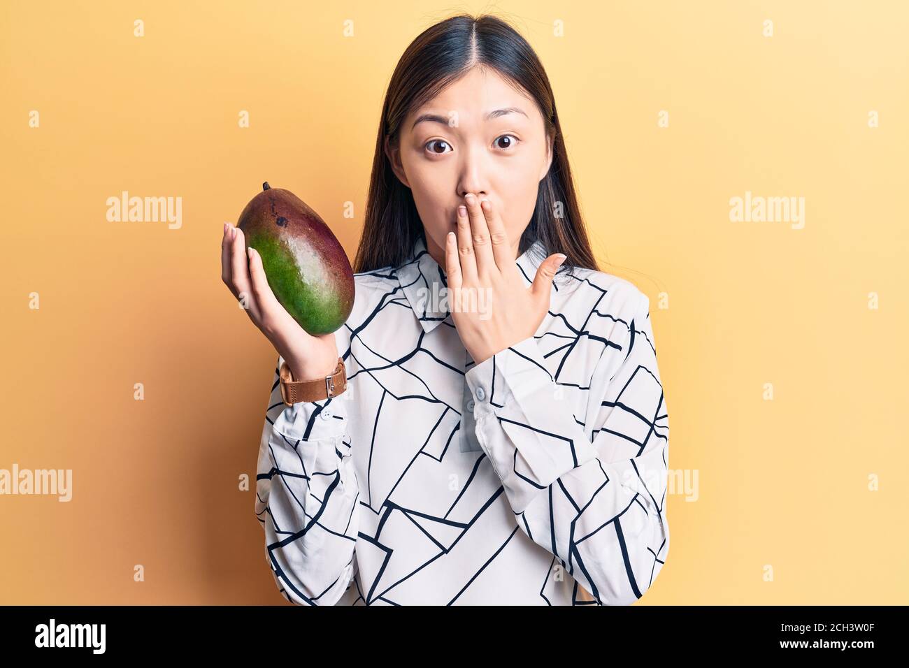 Young beautiful chinese woman holding mango covering mouth with hand ...