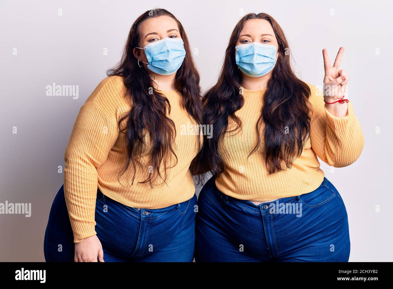 Young plus size twins wearing medical mask showing and pointing up with ...
