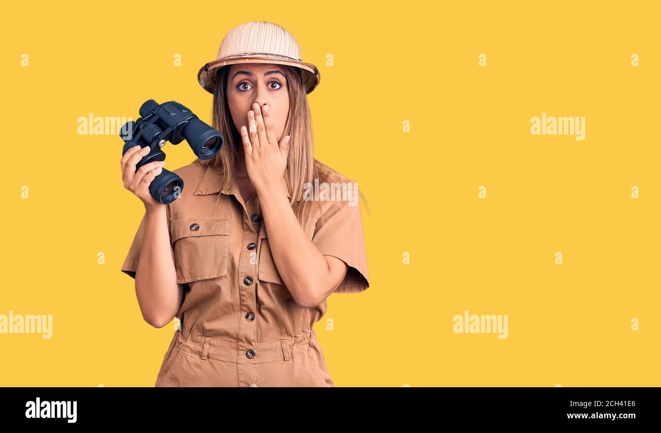 Young beautiful woman wearing explorer hat holding binoculars covering ...