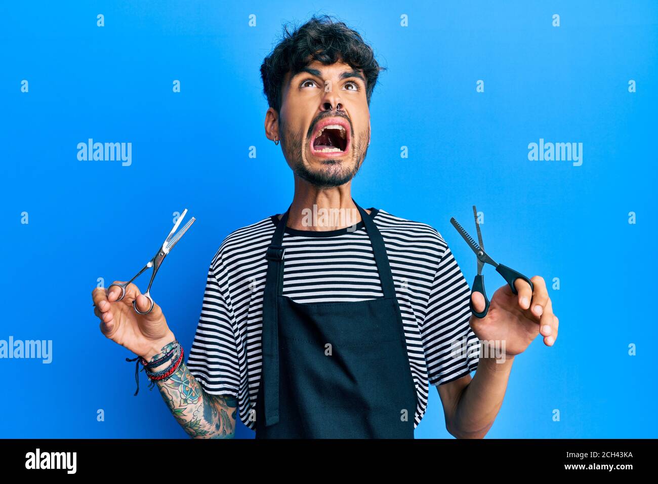 Young hispanic man wearing barber apron holding scissors angry and mad screaming frustrated and ...