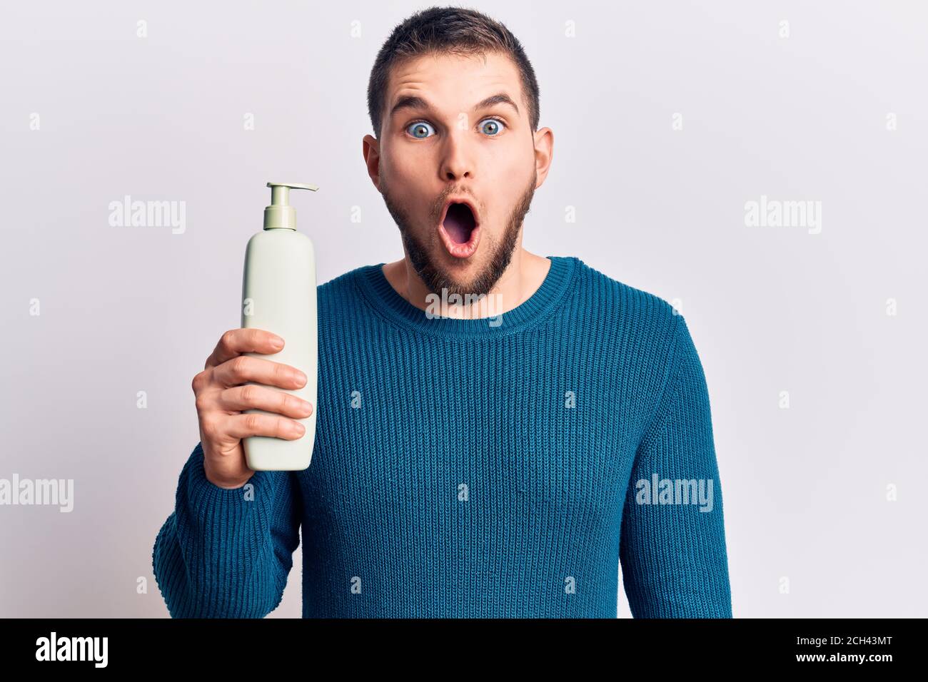 Young handsome man holding bottle of cream scared and amazed with open ...