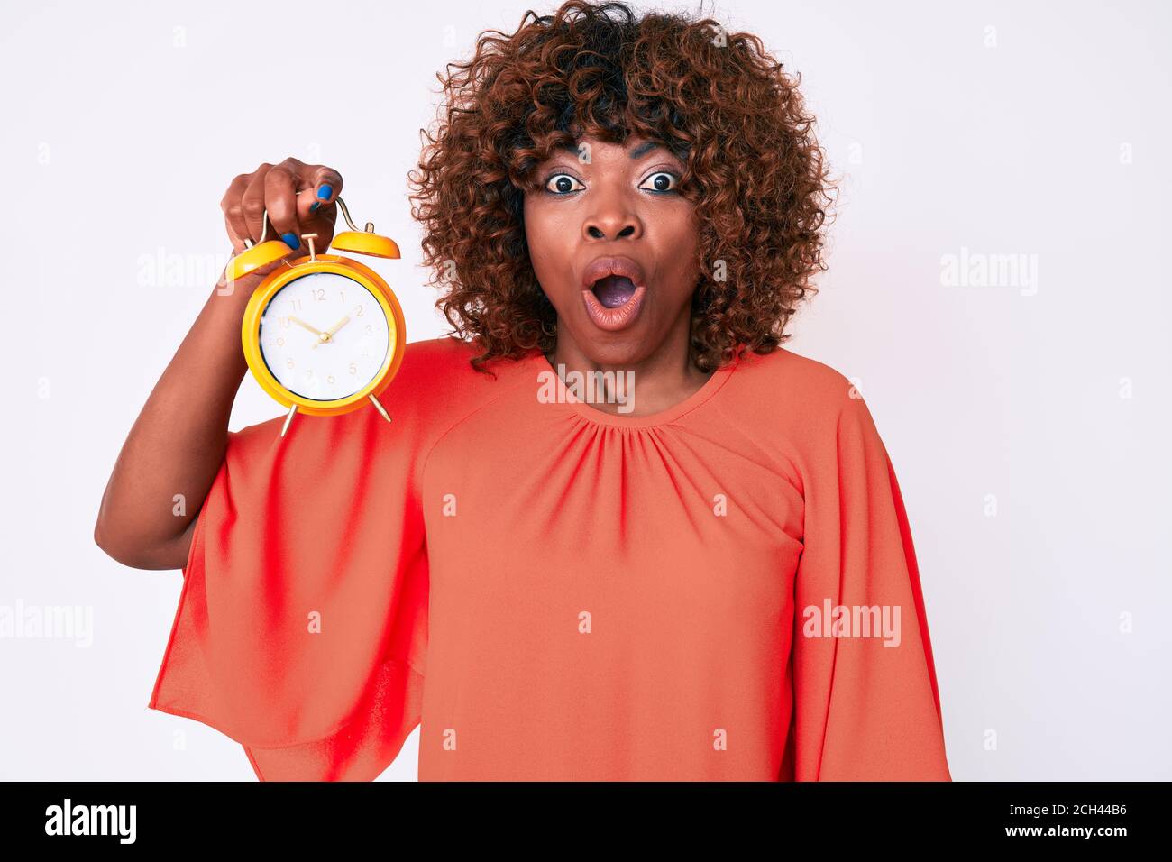 Young african american woman holding alarm clock scared and amazed with ...