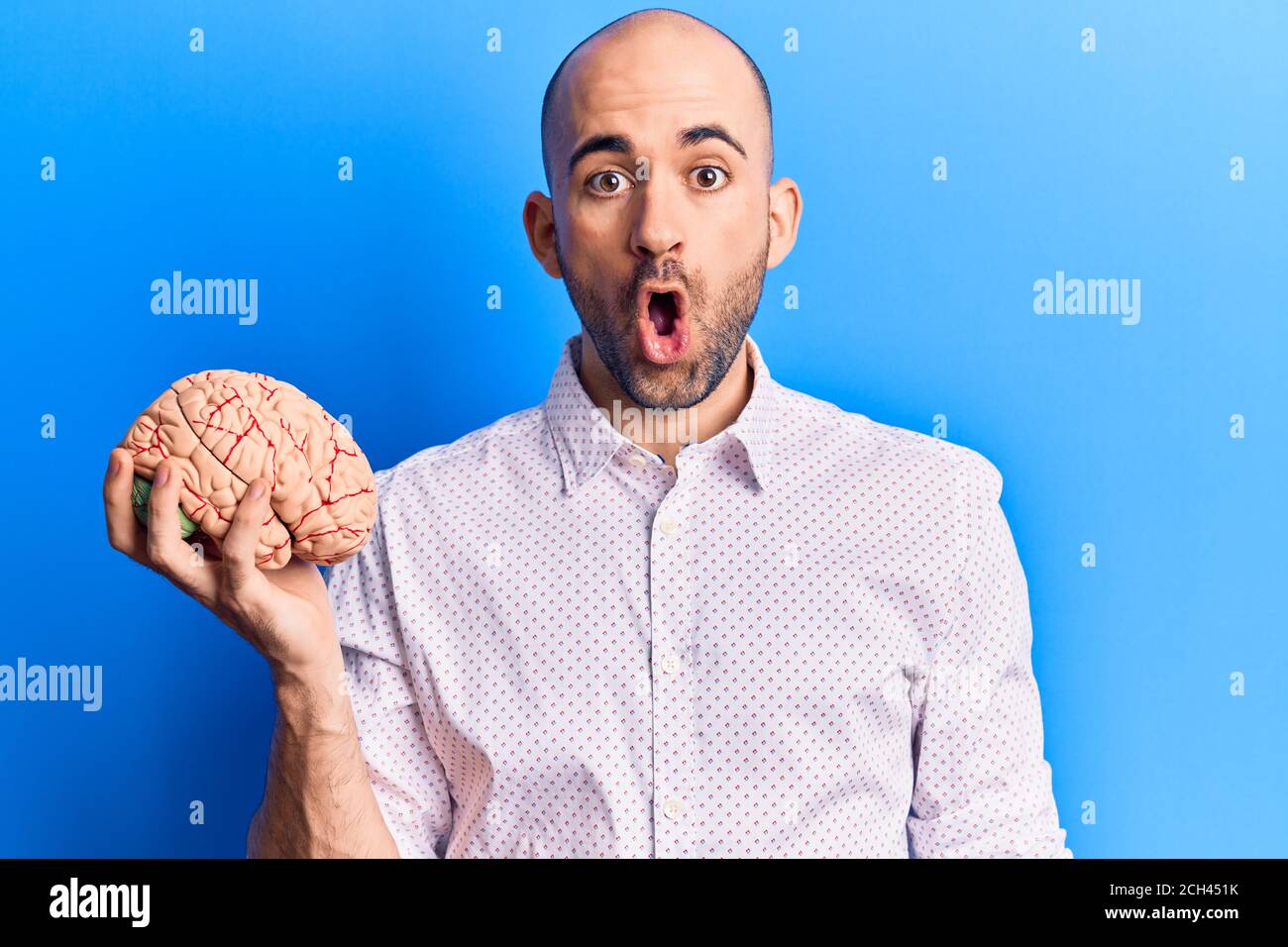 Young handsome bald man holding brain scared and amazed with open mouth ...