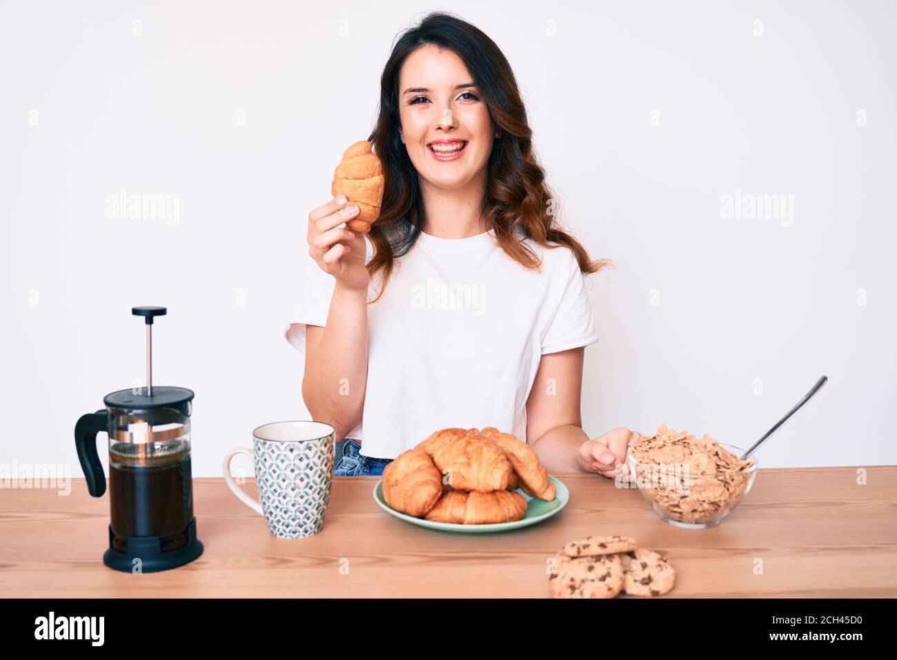 Young beautiful brunette woman eating breakfast holding croissant ...