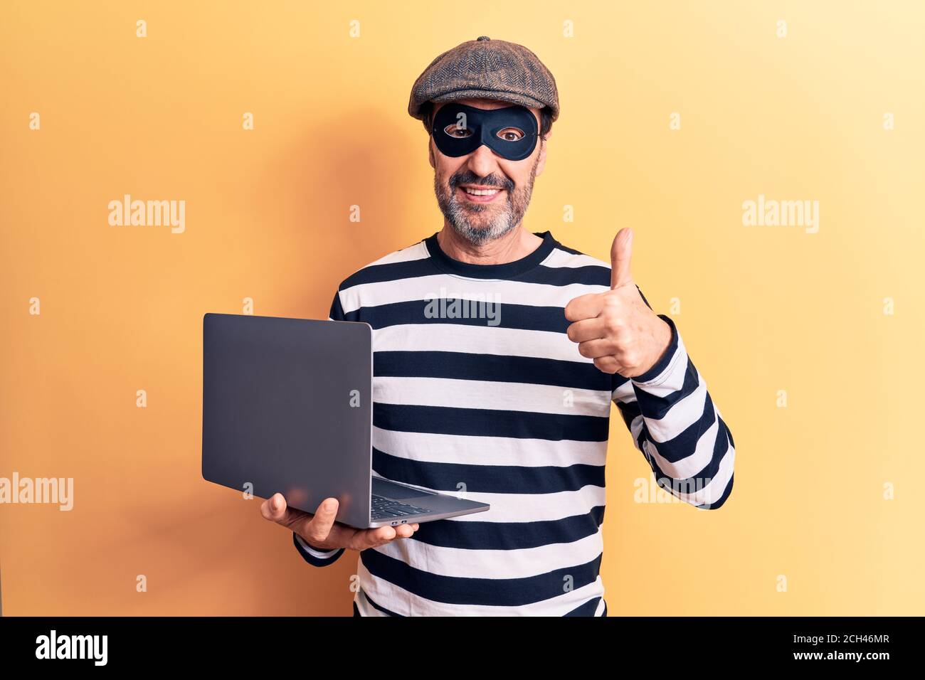 Middle age handsome burglar man wearing cap and mask using laptop over ...