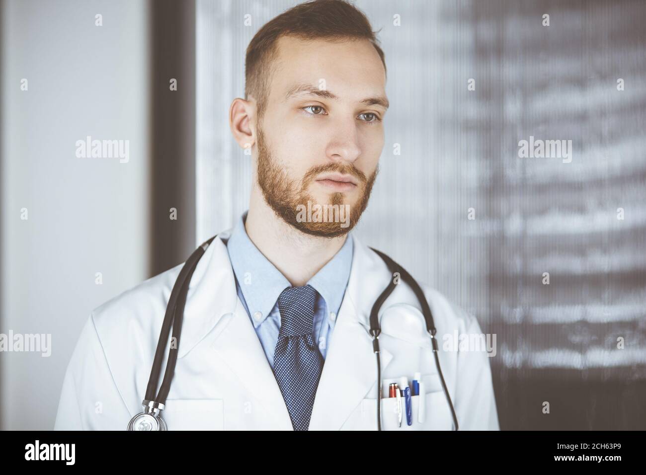 Red-bearded doctor standing straight in clinic near his working place ...