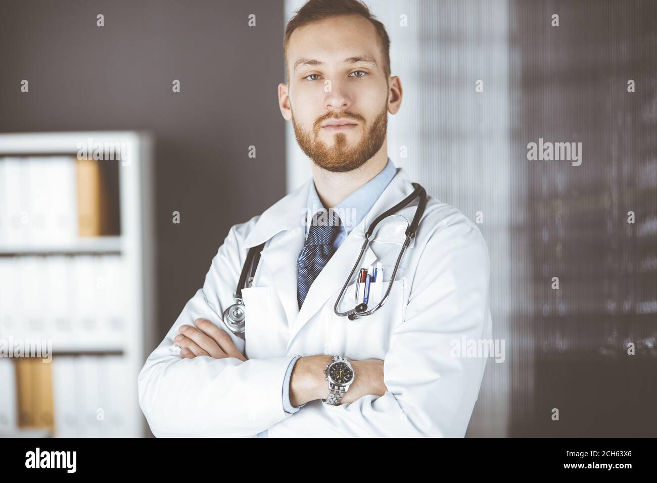 Red-bearded doctor standing straight in clinic near his working place ...