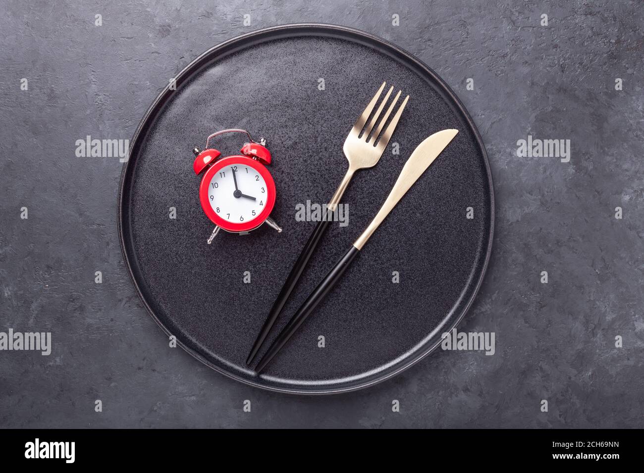 Red alarm clock, fork, knife and empty black ceramic plate on dark ...