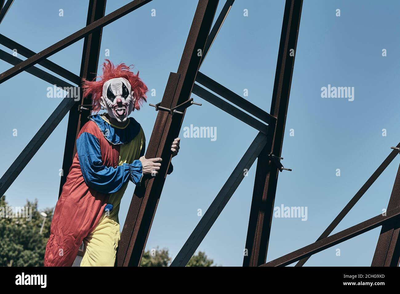 a scary evil clown, wearing a yellow, red and blue costume outdoors ...
