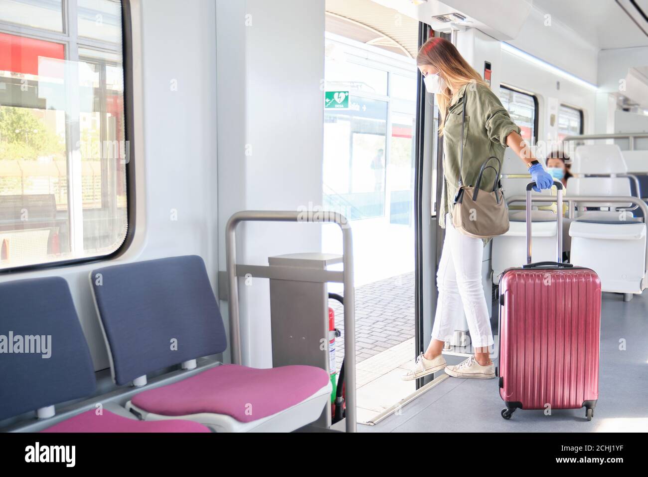 Young woman wearing face mask and gloves, carrying a suitcase getting ...