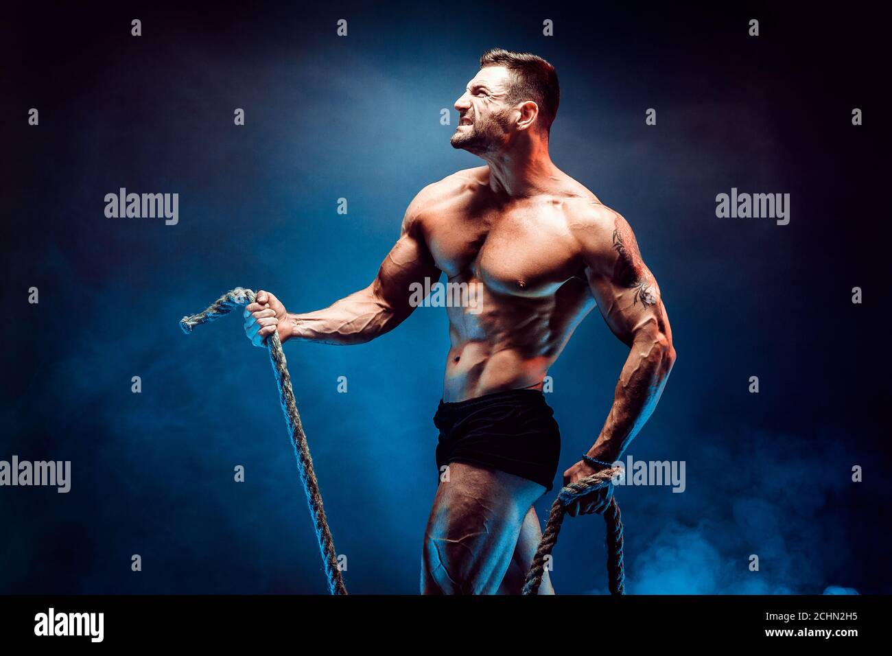 Side view of muscular bodybuilder man training hard with rope in studio ...