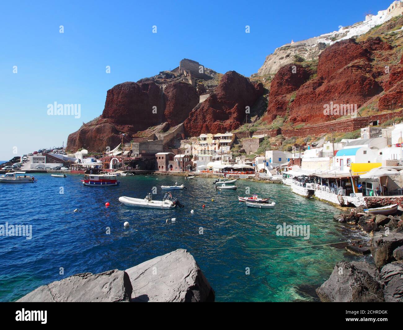 Boats moored in Amoudi Bay beneath a clear blue sky and against red ...