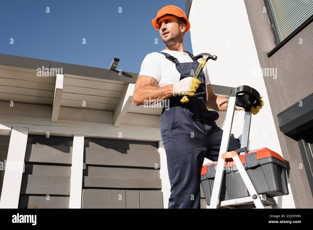 Builder in overalls and gloves holding hammer beside toolbox on ladder ...