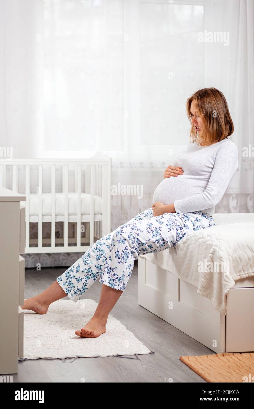 A young pregnant woman sits on the bed and holds her stomach, feeling ...