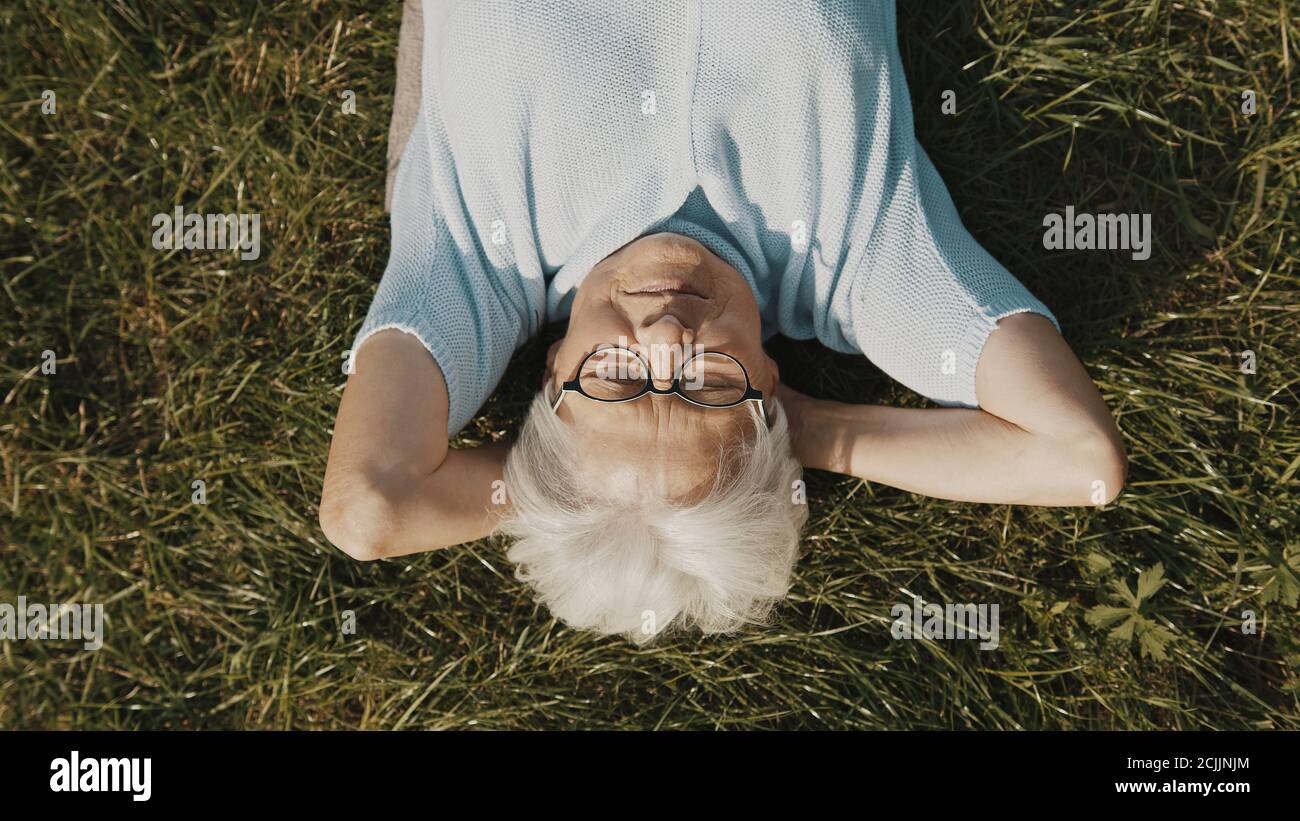 gray haired woman pensioner lying on the grass with hands under her ...
